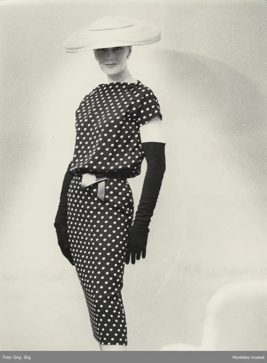 Modell i prickig klänning med skärp, vit hatt och operahandskar ...