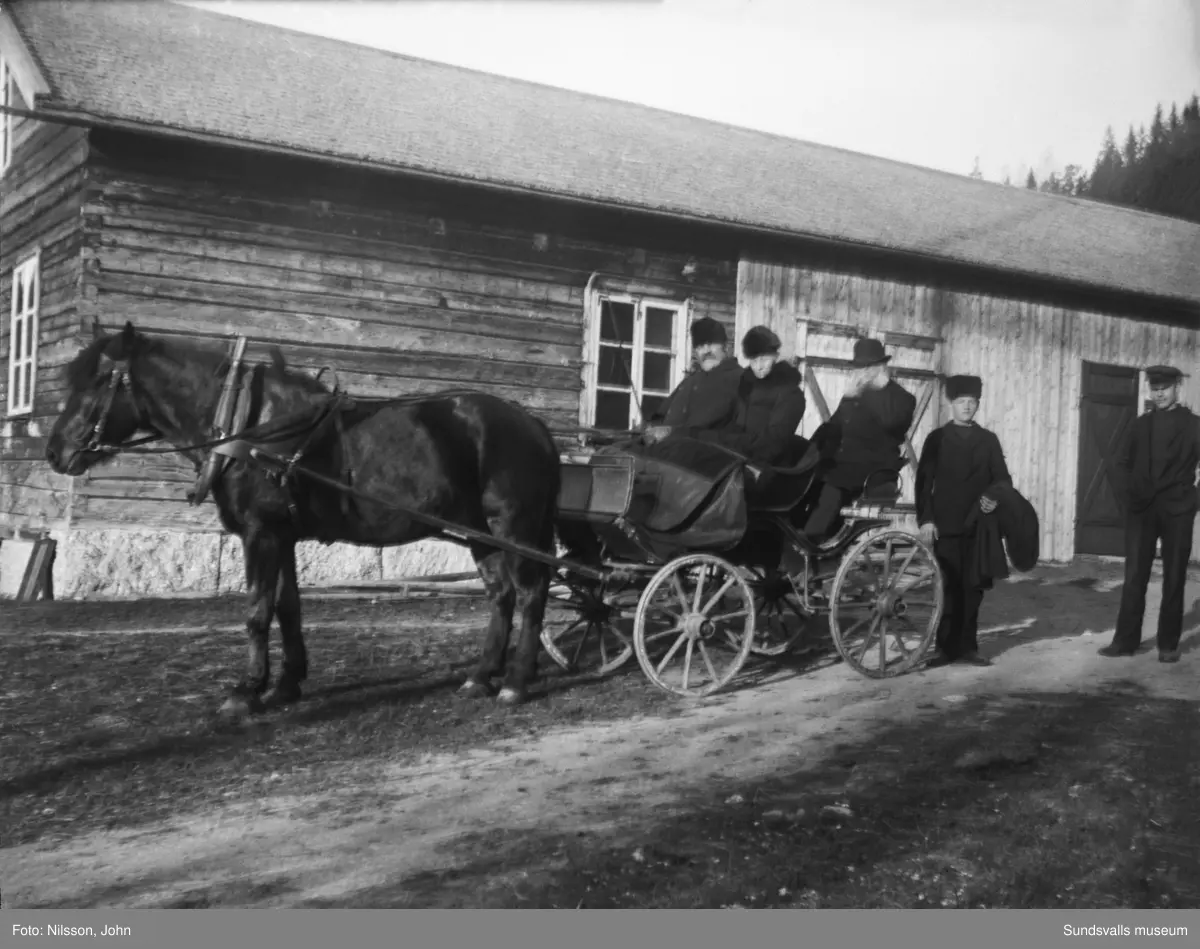 En häst spänd framför en fyrhjulig vagn intill ett timrat uthus ...