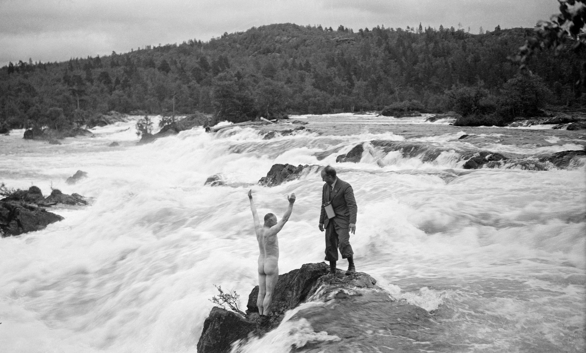 En naken mann står med armene strakt til værs på et skjær i Skogfossen (Reamal, Hakokoski) i ...