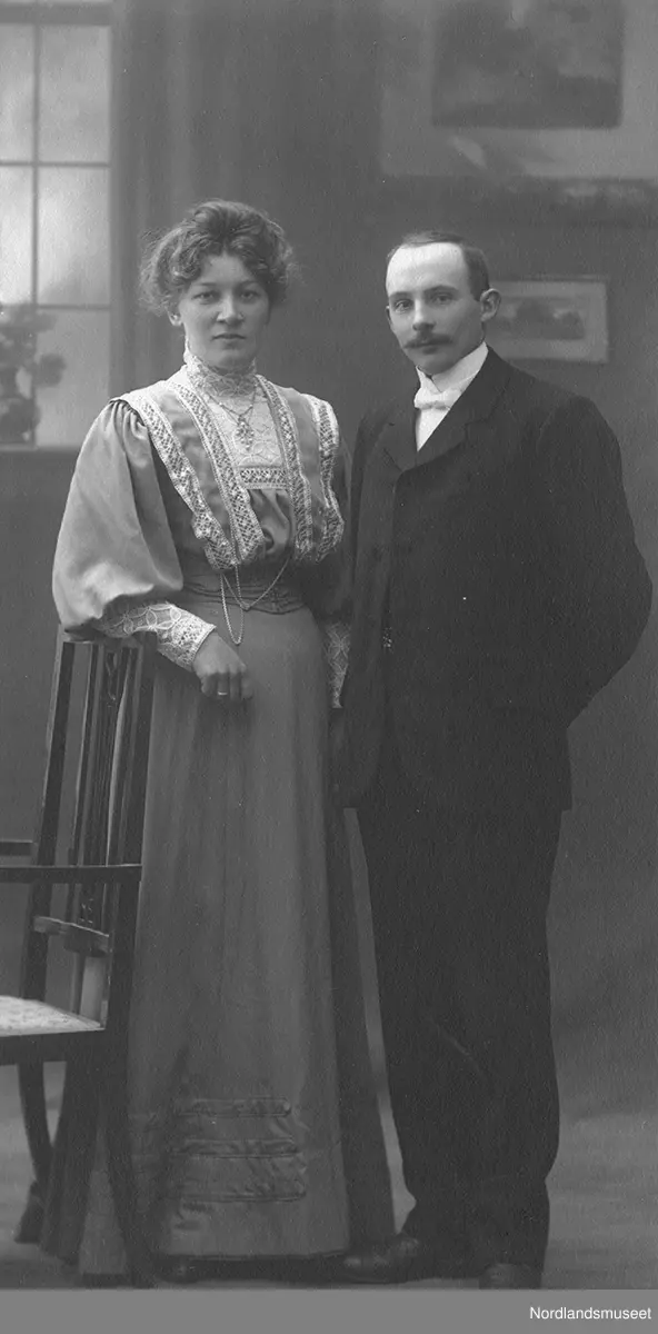 Portrett av et par stående i ei stue. - Nordlandsmuseet / DigitaltMuseum