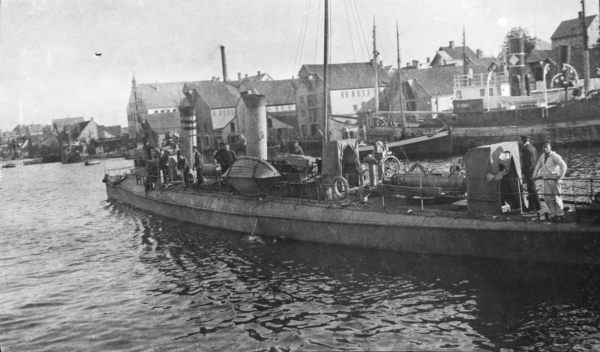 Gruppebilde. Lite dampskip med mannskap. Flere skip og sjøhusrekke i ...